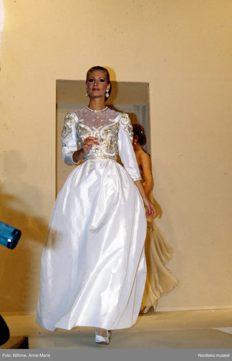 Modevisning. Modell i vit långklänning med pärlor och guldapplikationer ...