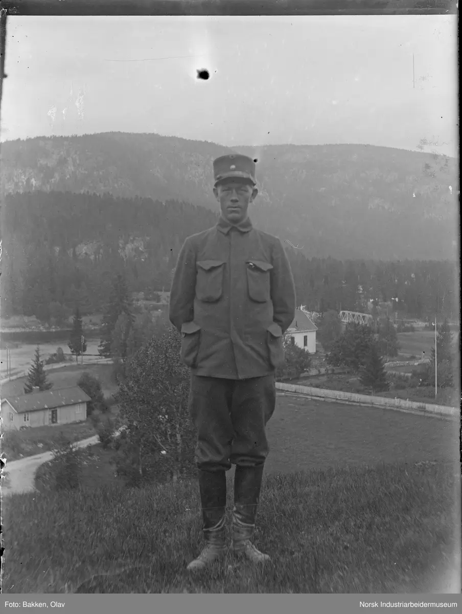 Mann i uniform stående utendørs. Gransherad Kirke i bakgrunnen. - Norsk ...