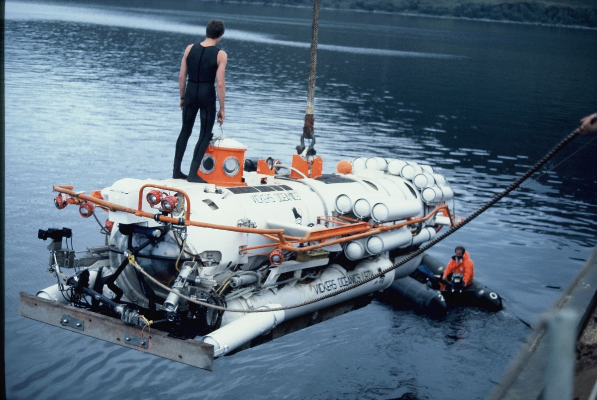 En ubåt løftes fra en lektert og ut i sjøen. En mann i våtdrakt står ...