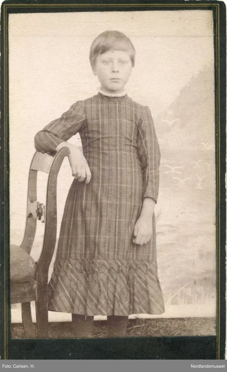 Portrettbilde av en jente i rutet kjole som lener seg til en stol ...