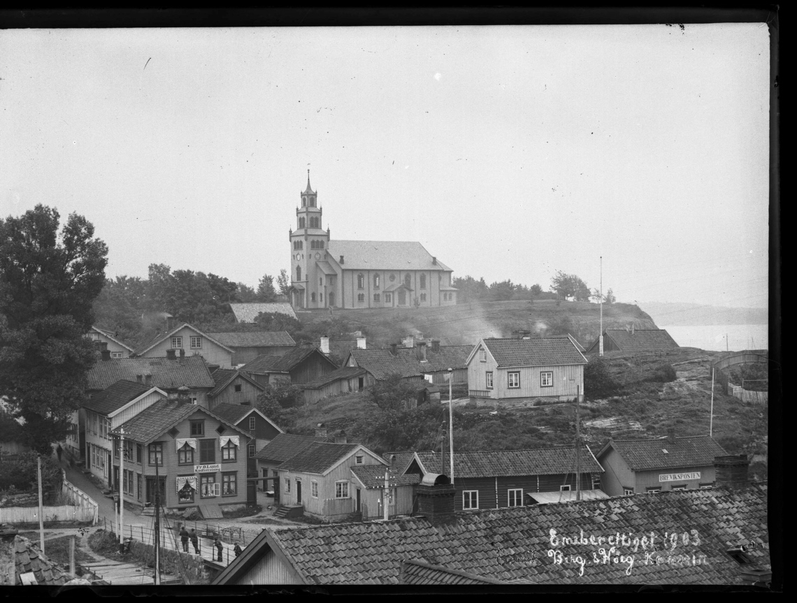 Gamle Brevik kirke med bebyggelse nedenfor. På en bro står det ...