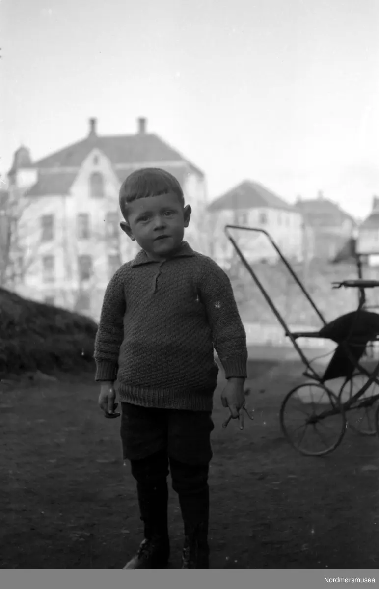 Et barn stående utendørs. En vogn til høyre i bildet. Bygninger i ...