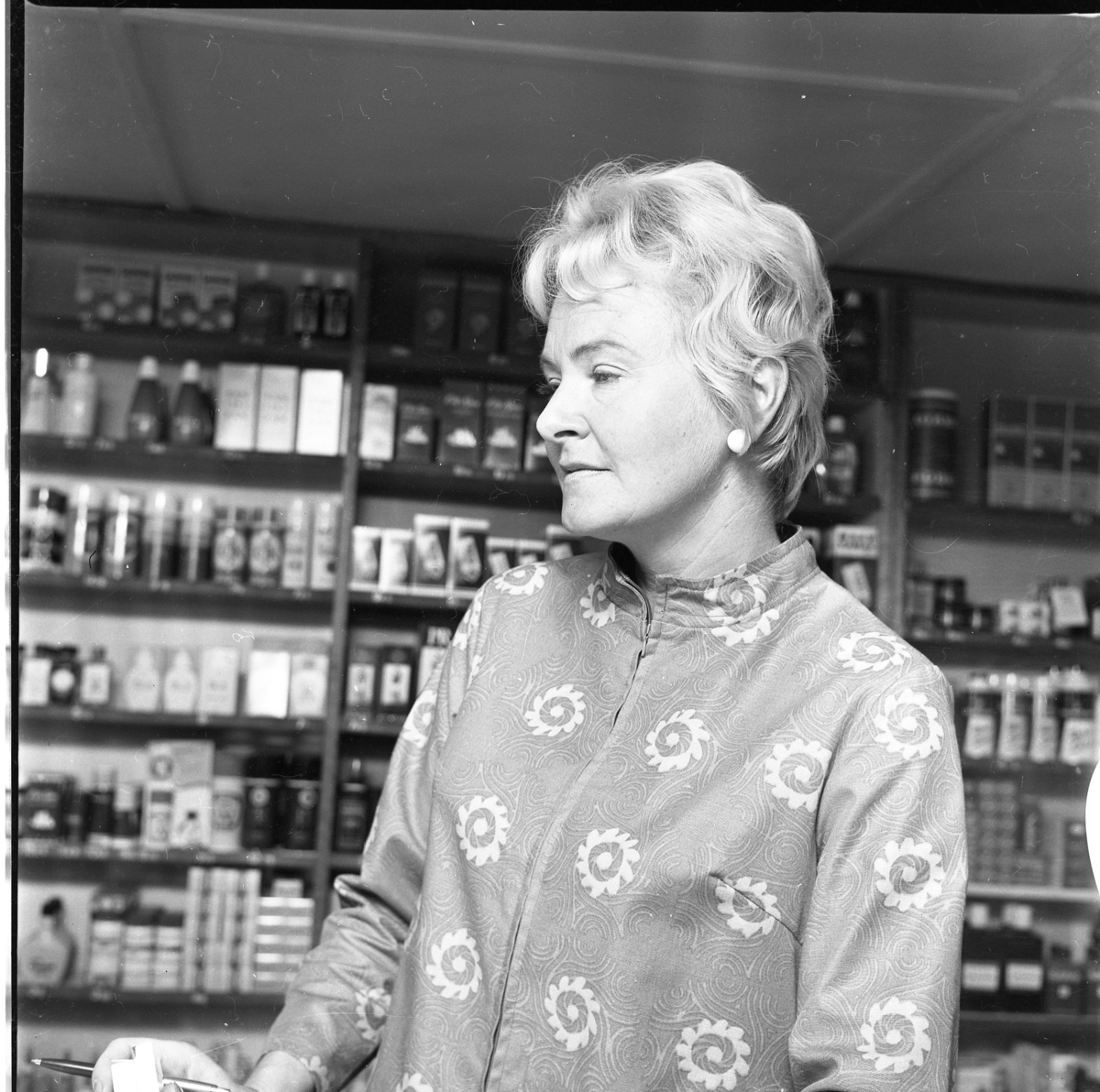 Birgit Roman står bakom disken i en butik. Vägghyllor med olika ...